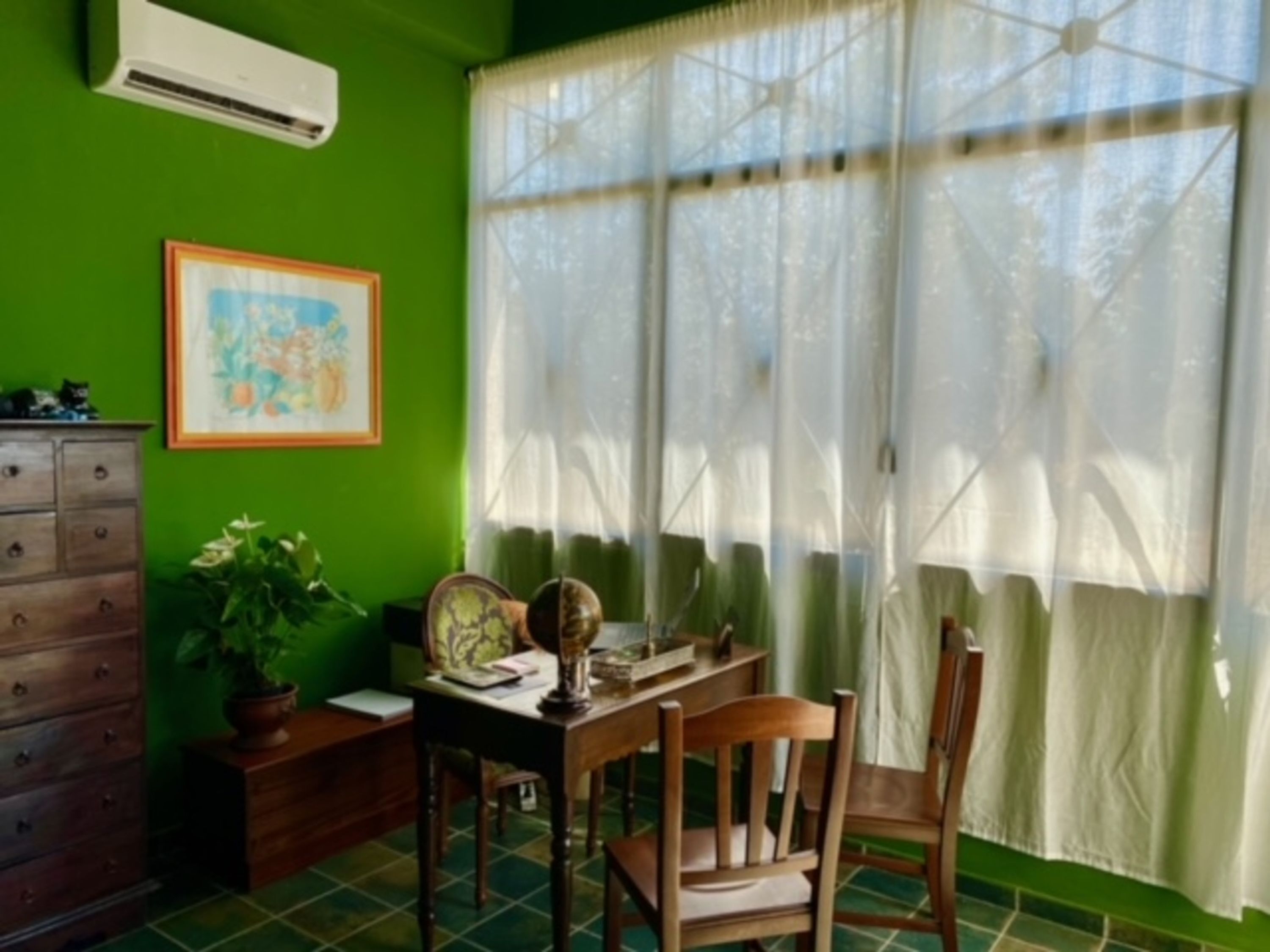 Quarto com parede verde, cómoda de madeira, escrivaninha com cadeira, planta em vaso e janela com cortinas leves.