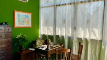 Quarto com parede verde, cómoda de madeira, escrivaninha com cadeira, planta em vaso e janela com cortinas leves.