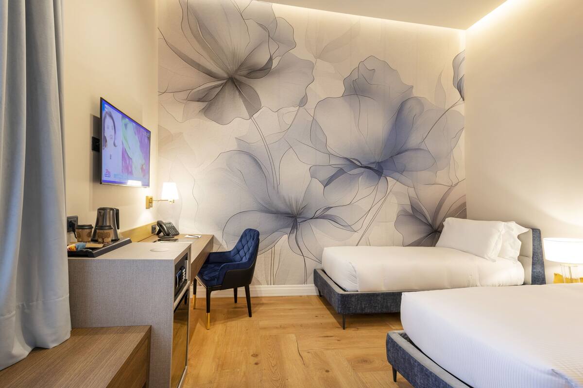 Quarto de hotel com papel de parede floral, cama, secretária com cadeira, máquina de café e televisão.