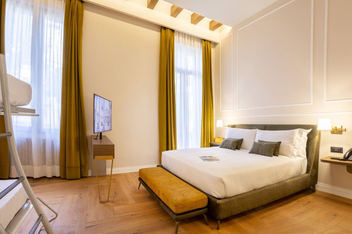 Quarto de hotel bem arrumado com uma cama grande, mesa de cabeceira, um banco e uma televisão sobre suporte.