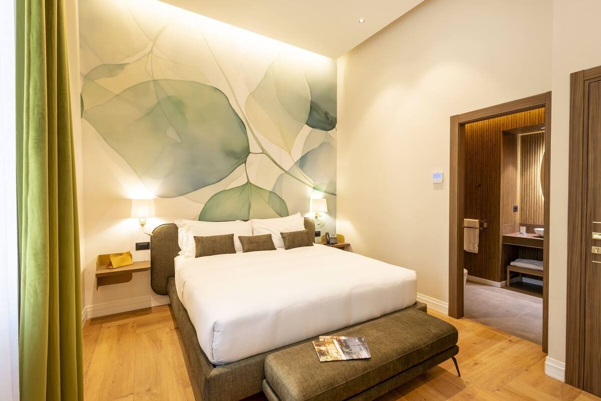 Quarto de hotel moderno com uma cama grande, um banco, um mural na parede e uma porta de madeira.