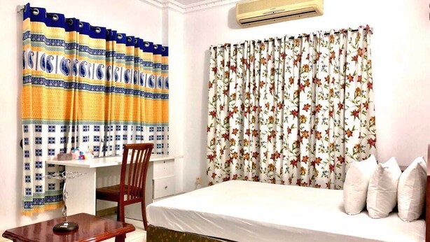 Quarto com cama, escrivaninha e cadeira. Há uma janela com cortinas e um ar condicionado acima da cama.