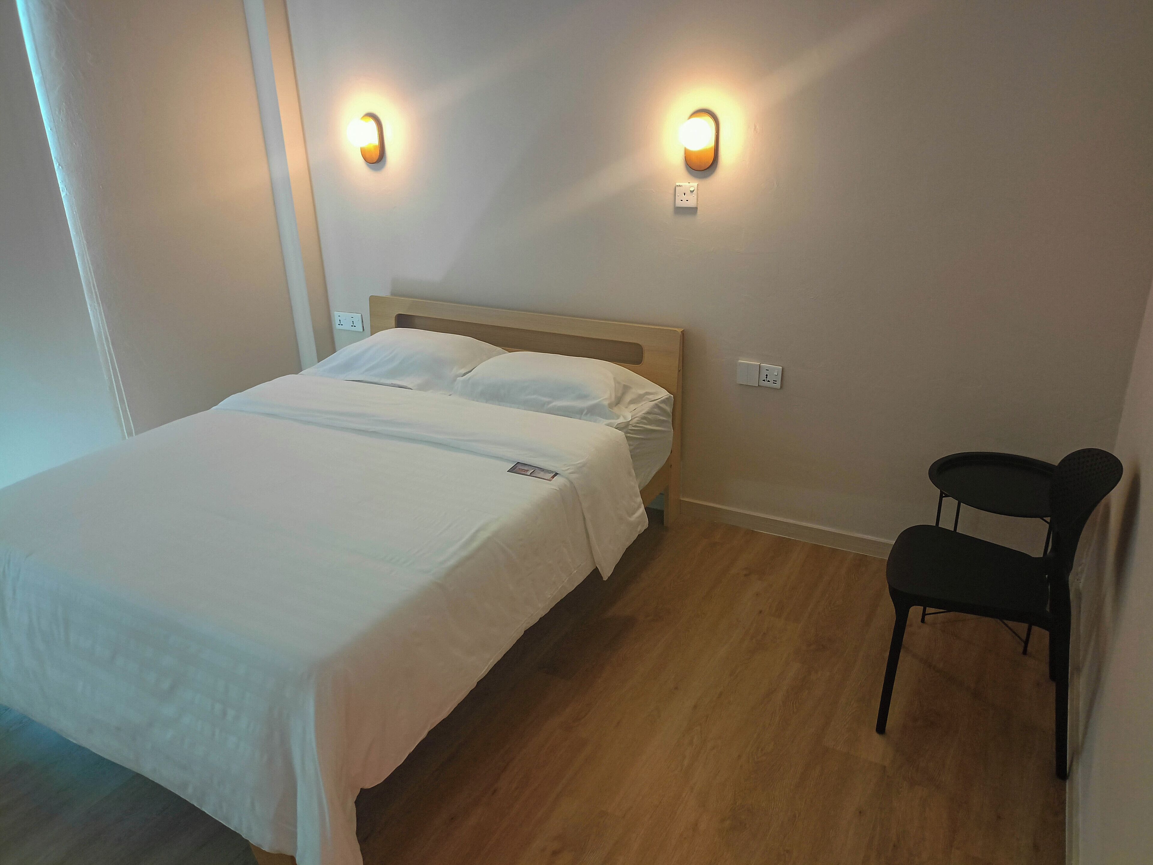 Quarto de hotel com cama, duas luminárias fixadas na parede, uma tomada e uma cadeira preta.