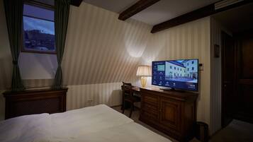 Quarto de hotel com cama, escrivaninha com luminária, televisão exibindo vista urbana e janela com cortinas.