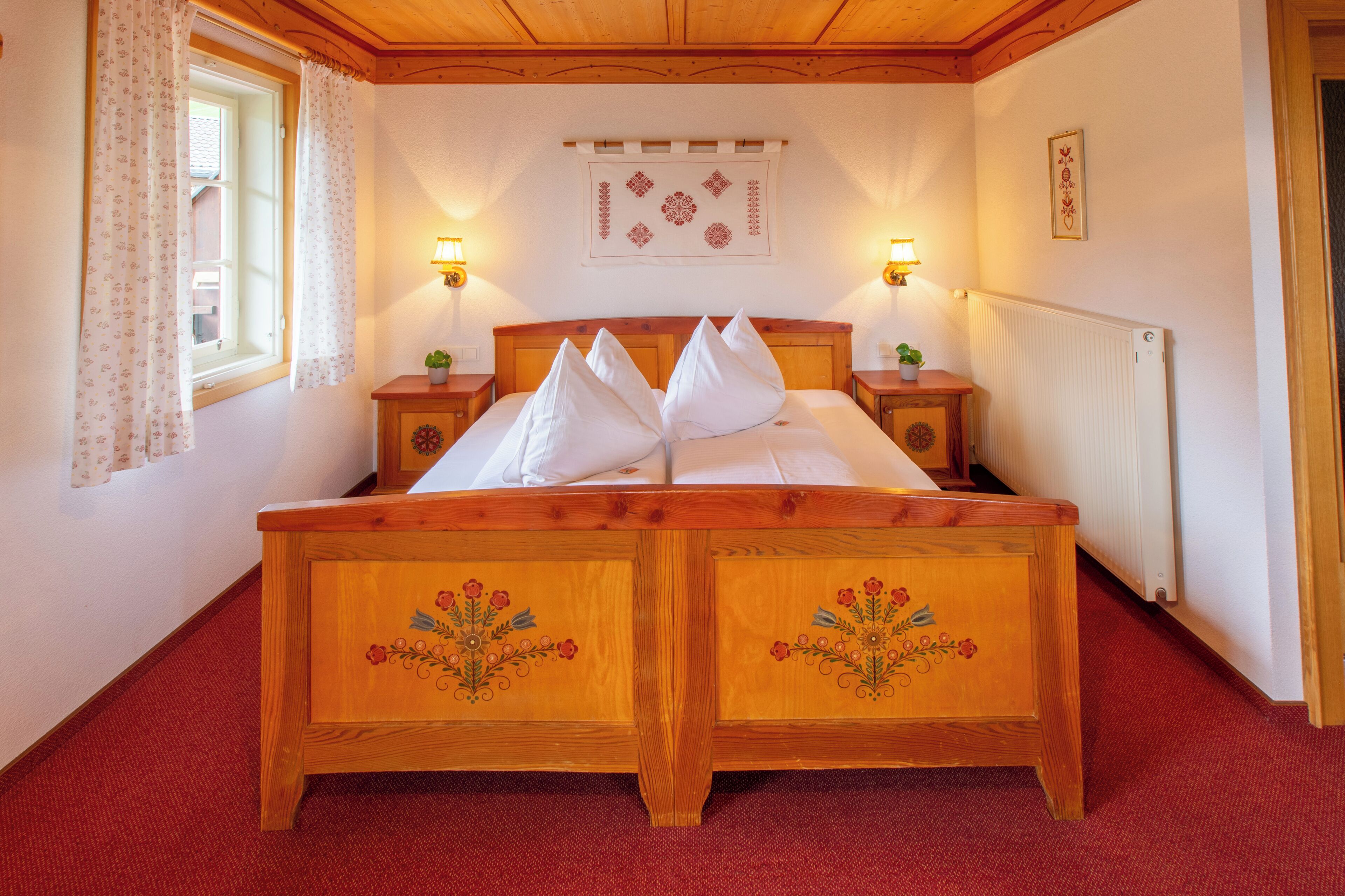 Uma cama de madeira com detalhes entalhados em flor, duas mesas de cabeceira com luminárias, uma janela com cortinas e um tapeçaria na parede.