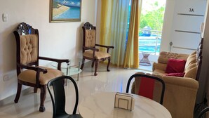 Uma sala de estar com ventilador de teto, um quadro, sofá, mesa com cadeiras e vista para o exterior pela janela.