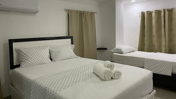 Quarto de hotel com uma cama de solteiro e uma cama menor, ambas com roupa de cama branca e colchas estampadas. Há duas cortinas bege, uma em cada janela.