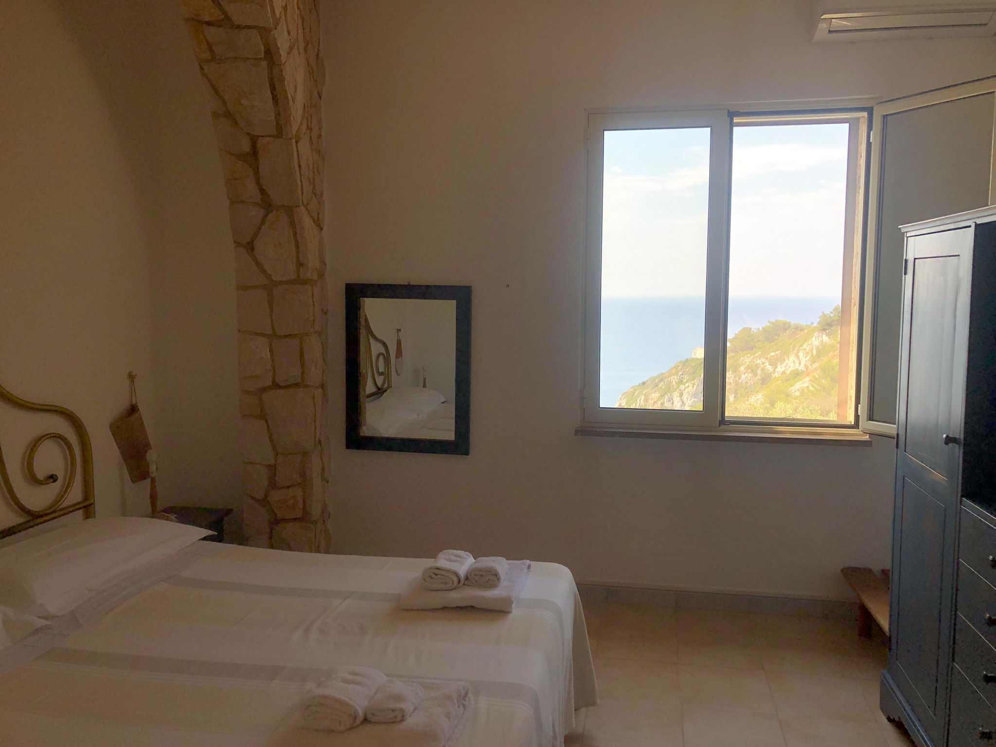 Um quarto com parede de pedra, cama com roupa de cama branca, um quadro na parede e uma janela com vista para o mar.