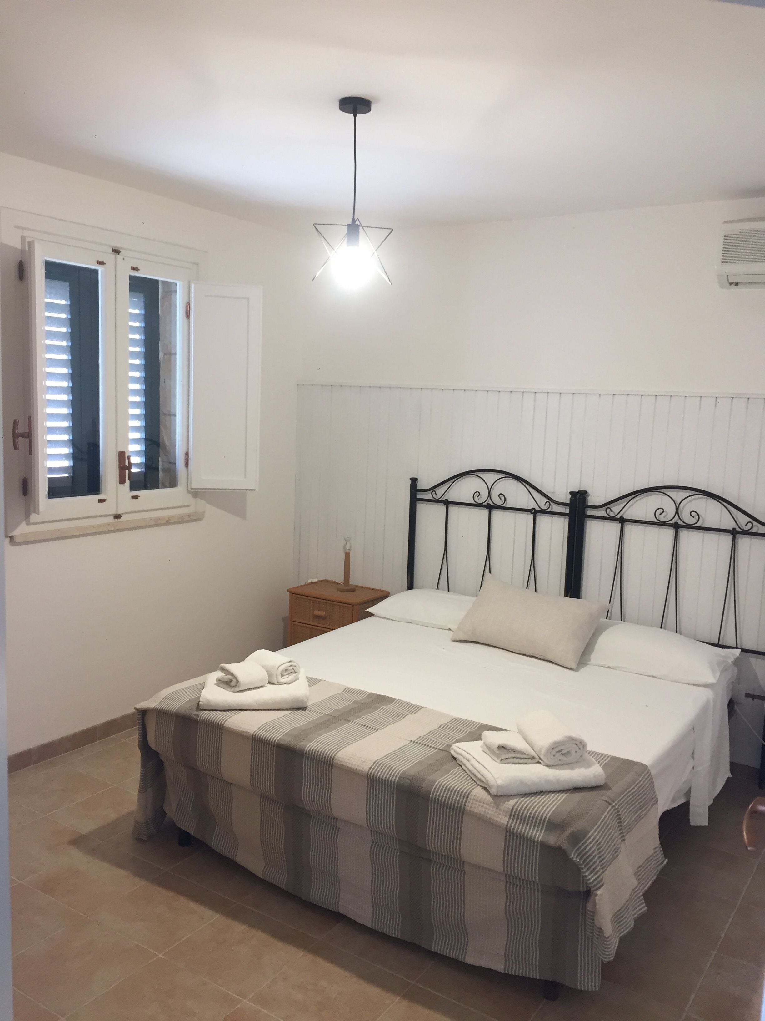 Um quarto com cama, mesa de cabeceira, persianas na janela e luminária no teto.