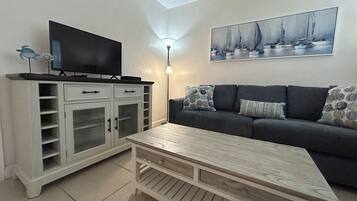 Uma sala de estar com uma TV sobre um móvel, um sofá com almofadas estampadas e um quadro de veleiros na parede.