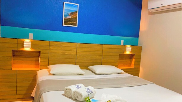 Quarto de hotel com cabeceira de madeira, duas almofadas, cama bem arrumada com roupa de cama branca e fotografia de paisagem costeira na parede.