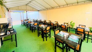Uma área de refeições coberta por tendas, com mesas e cadeiras, um espaço para churrasco e plantas em vasos.
