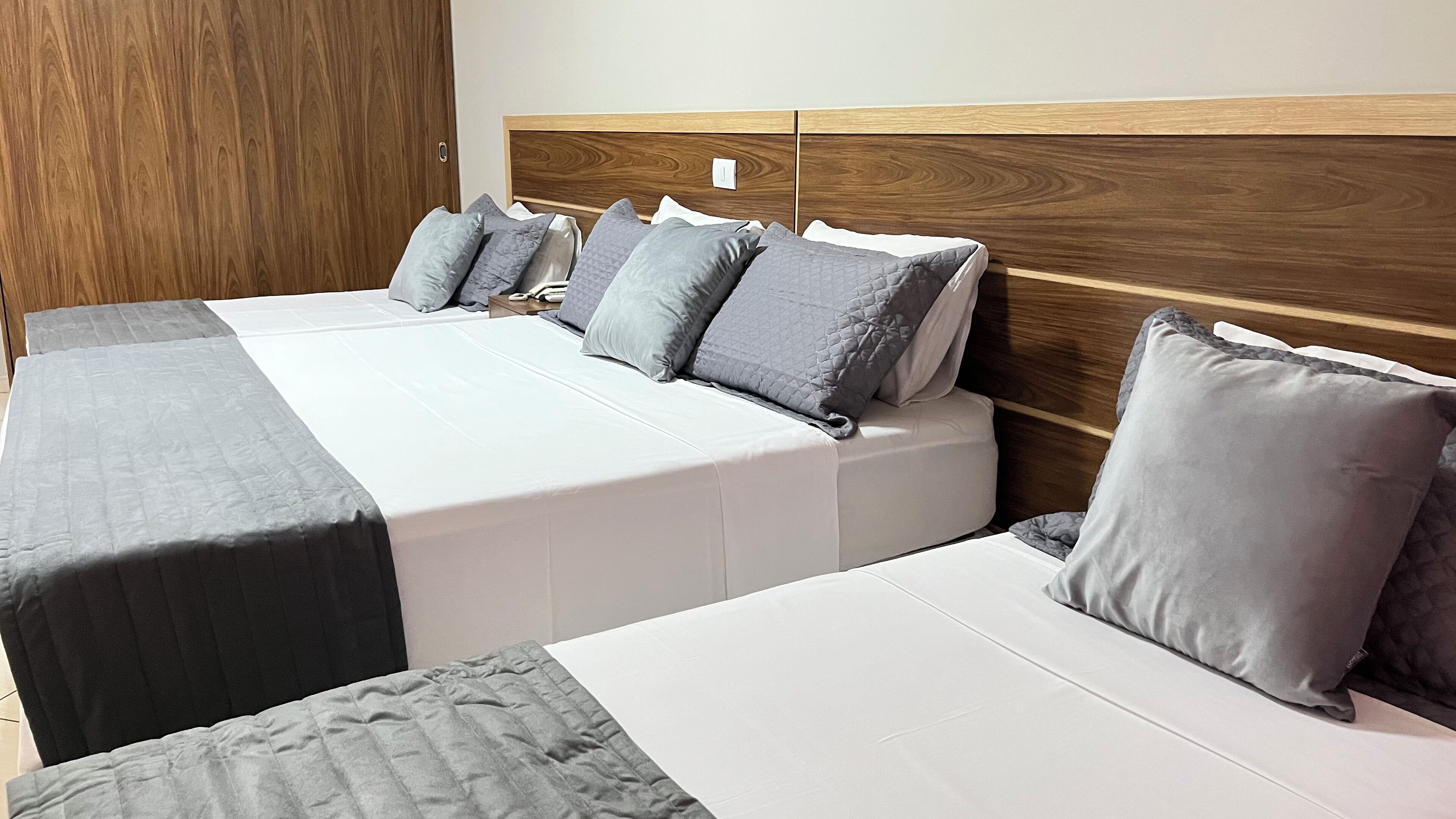 Uma cama de casal com cabeceira de madeira, roupa de cama cinzenta e várias almofadas.