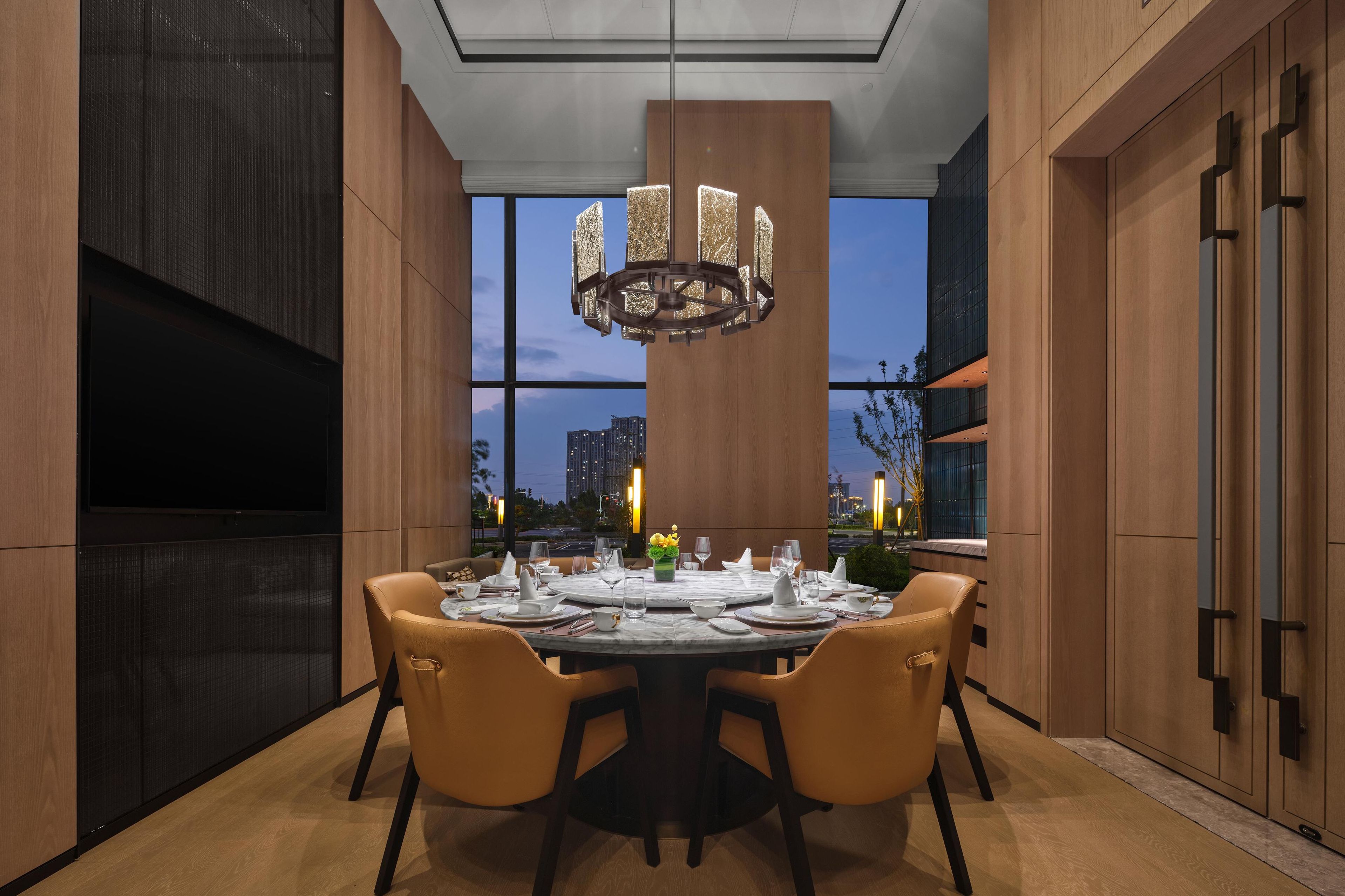 Uma área de jantar moderna com uma mesa redonda posta para seis pessoas, rodeada por cadeiras de couro. Um grande candelabro pendurado no teto e o espaço possui grandes janelas com vista para a cidade.