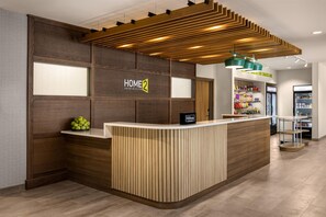 Bancada de recepção de hotel com design moderno, apresentando área de atendimento em madeira, sinalização com 'HOME 2 Suites by Hilton' e um bar de lanches ao fundo.