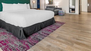 Quarto de hotel moderno com uma cama grande, uma escrivaninha e uma cadeira. Há um espelho, um abajur e um tapete pequeno.