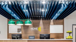 Uma moderna receção de hotel com design em painel de madeira, dois computadores HP e um letreiro indicando 'HOME 2 Suites by Hilton'.