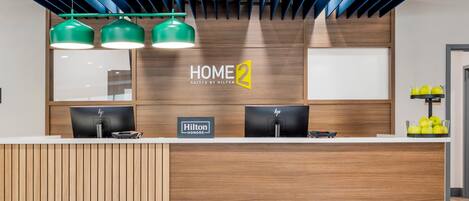 Uma moderna receção de hotel com design em painel de madeira, dois computadores HP e um letreiro indicando 'HOME 2 Suites by Hilton'.