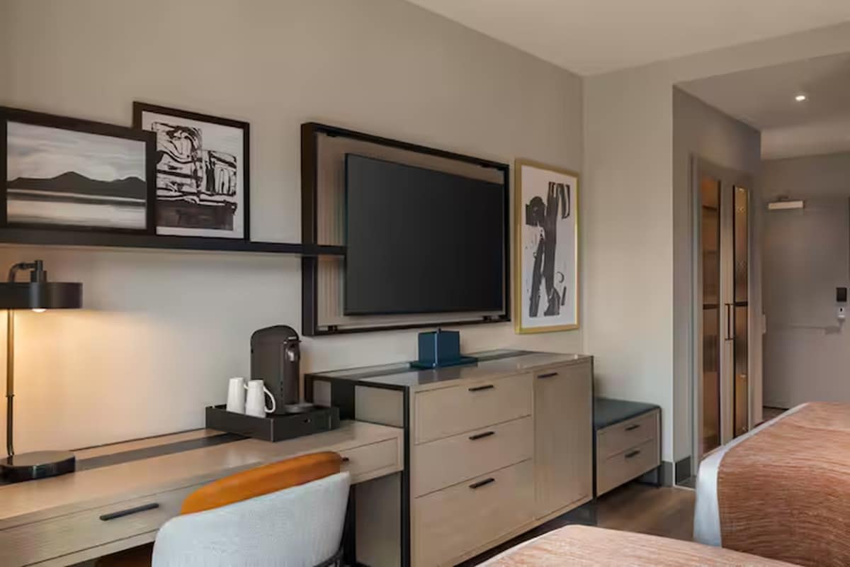 Quarto de hotel moderno com uma televisão de tela plana grande fixada na parede, uma escrivaninha com luminária e uma cama com cabeceira de madeira.