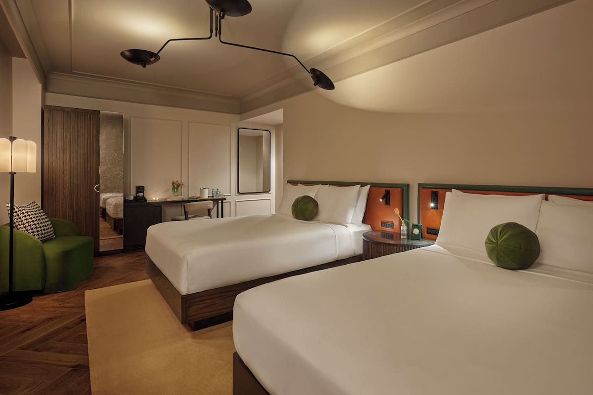 Quarto de hotel com duas camas, uma escrivaninha com cadeira, um espelho e uma poltrona verde.