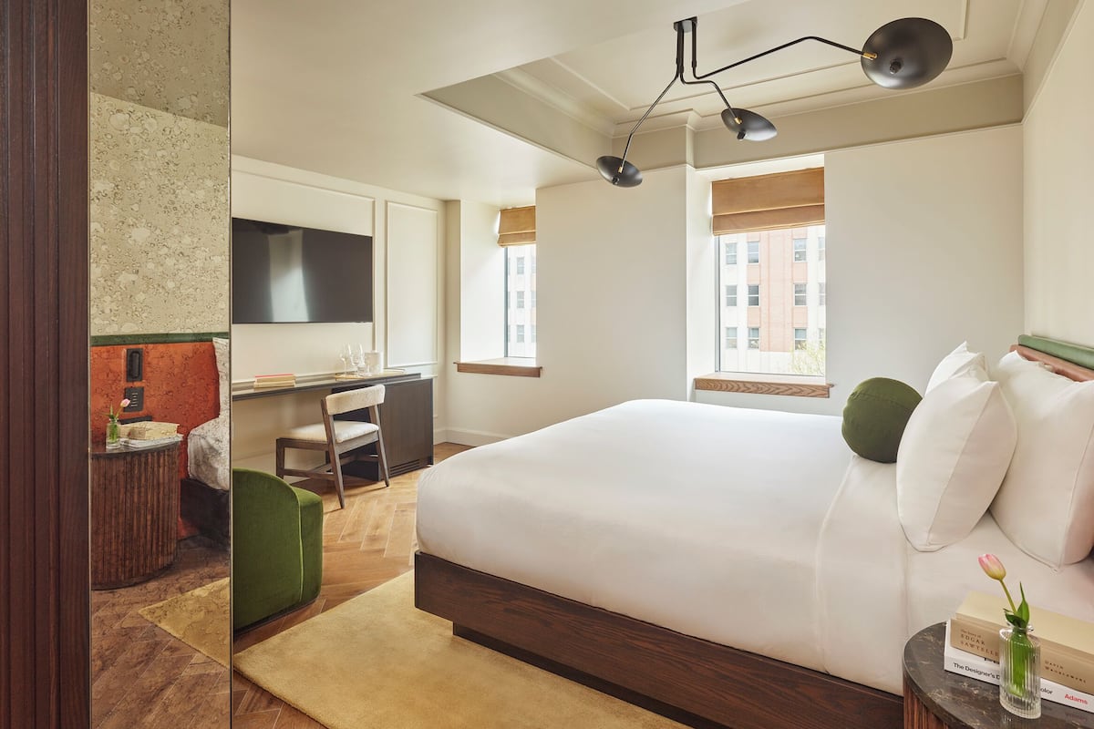 Quarto de hotel moderno com uma cama grande, uma escrivaninha e uma cadeira. Há uma janela com vista para edifícios.