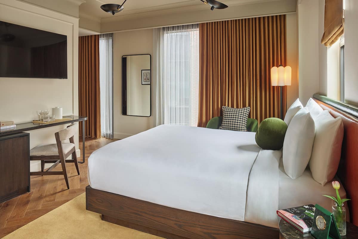 Quarto de hotel com uma cama grande, uma escrivaninha com cadeira, televisão de tela plana, espelho e janela com cortinas.
