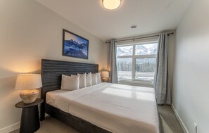 Quarto de hotel com uma cama grande, duas luminárias de cabeceira, um quadro na parede e uma janela com cortinas.