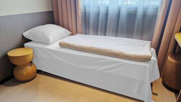 Uma cama de solteiro com colchão e travesseiro brancos, uma mesinha de cabeceira de madeira e uma janela com cortinas leves.