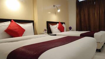 Quarto de hotel com duas camas, cada uma com roupa de cama branca e almofadas vermelhas, uma mesinha de cabeceira com abajur e cortina na janela.