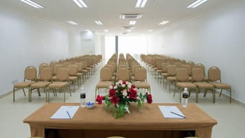 Uma sala de conferências com fileiras de cadeiras, uma mesa comprida com arranjos florais e uma tela de projetor na frente.