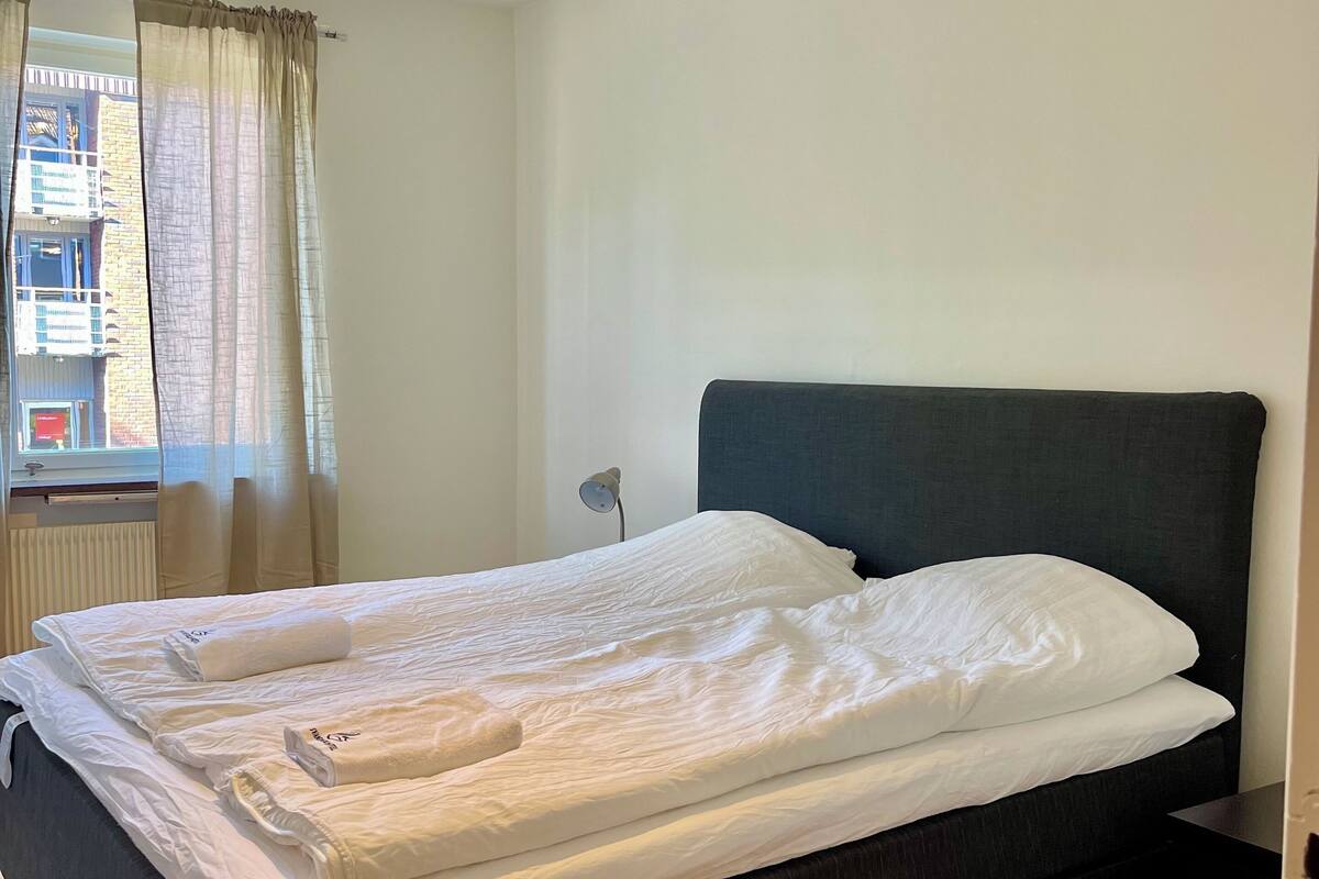 Cama bem arrumada com lençóis e toalhas brancas, uma cabeceira escura e um lustre no teto.