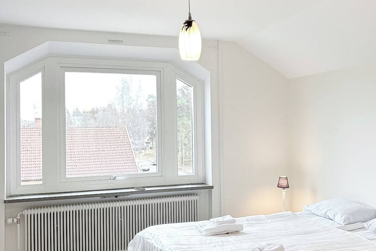 Quarto com uma janela grande, um radiador, uma cama com roupa de cama branca e um candeeiro suspenso.