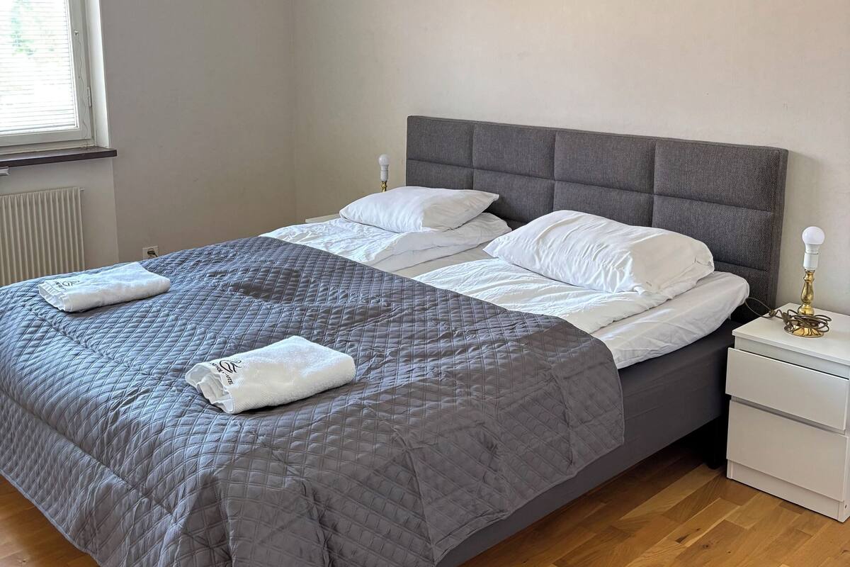 Cama bem arrumada com colcha cinza, almofadas brancas e abajur em uma mesinha de cabeceira branca.