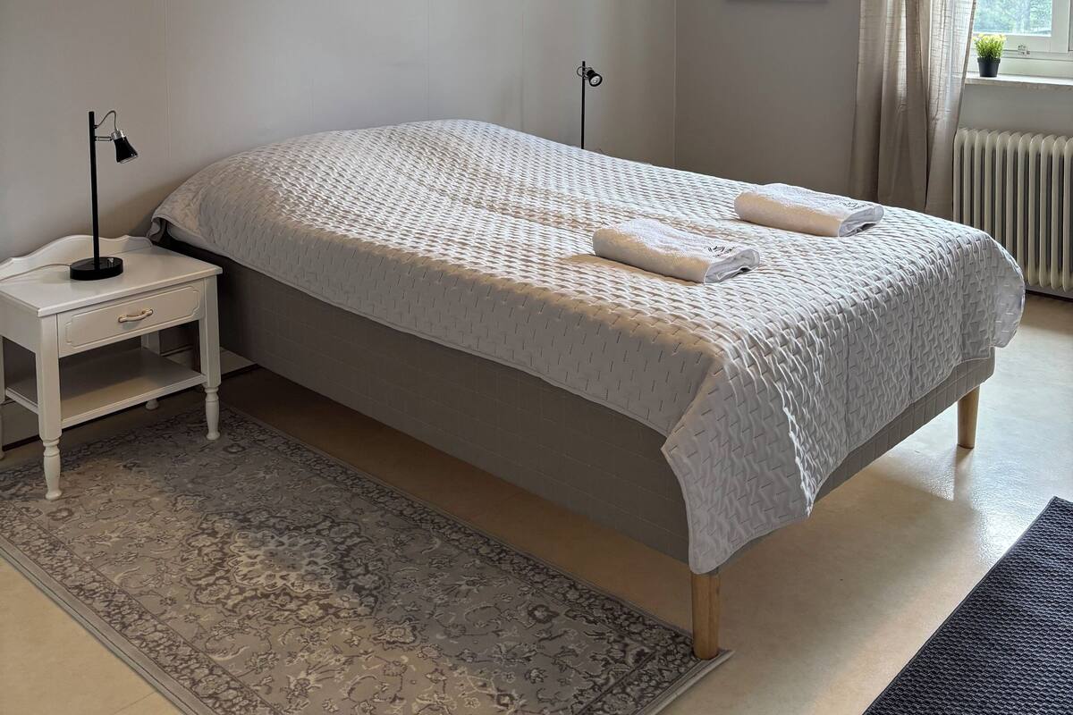 Quarto com cama, mesa de cabeceira, abajur, tapete e janela com cortinas.