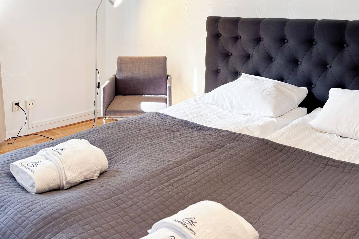 Cama bem arrumada com lençóis brancos e colcha cinza, cabeceira estofada escura, luminária de chão moderna e poltrona cinza.