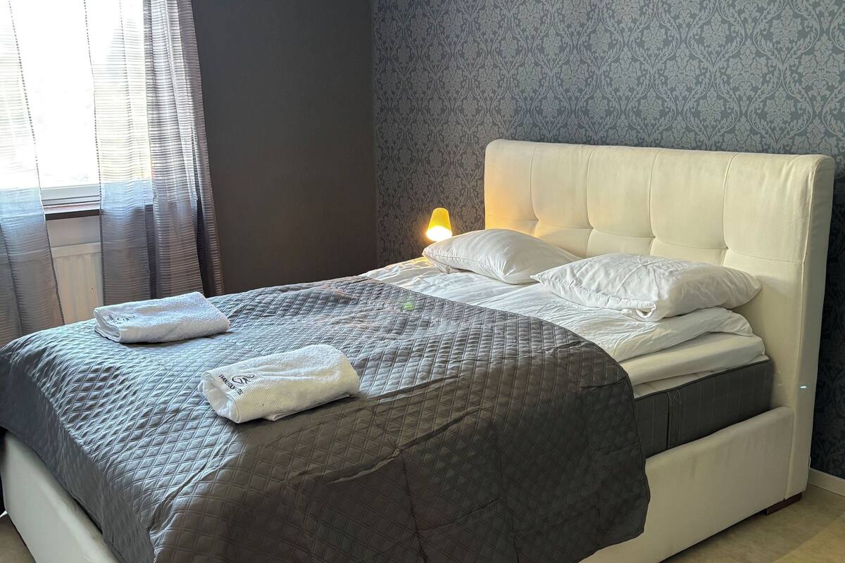 Uma cama bem arrumada com um espalha-cama cinza acolchoado, almofadas brancas e duas toalhas dobradas.