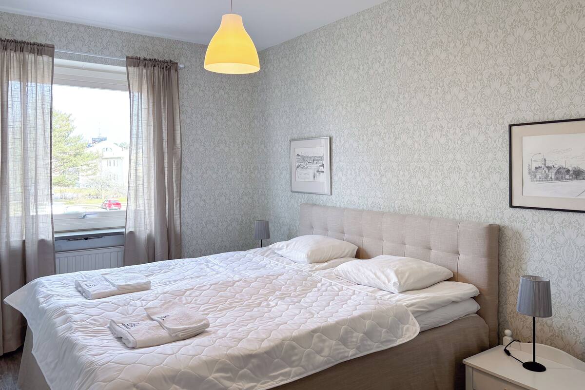 Uma cama bem arrumada com lençóis brancos e edredom acolchoado, um abajur e um quadro na parede.