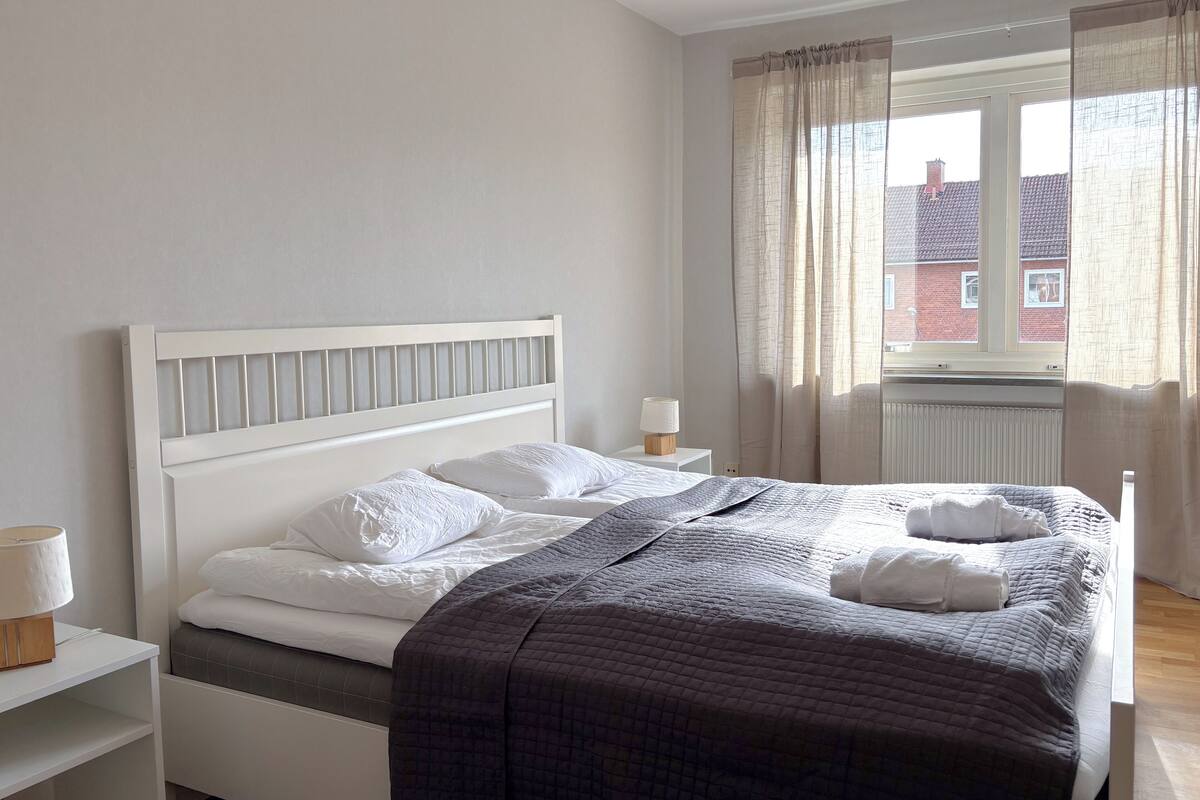 Cama bem arrumada com roupa de cama branca e edredom escuro, mesa de cabeceira com abajur e janela com cortinas.