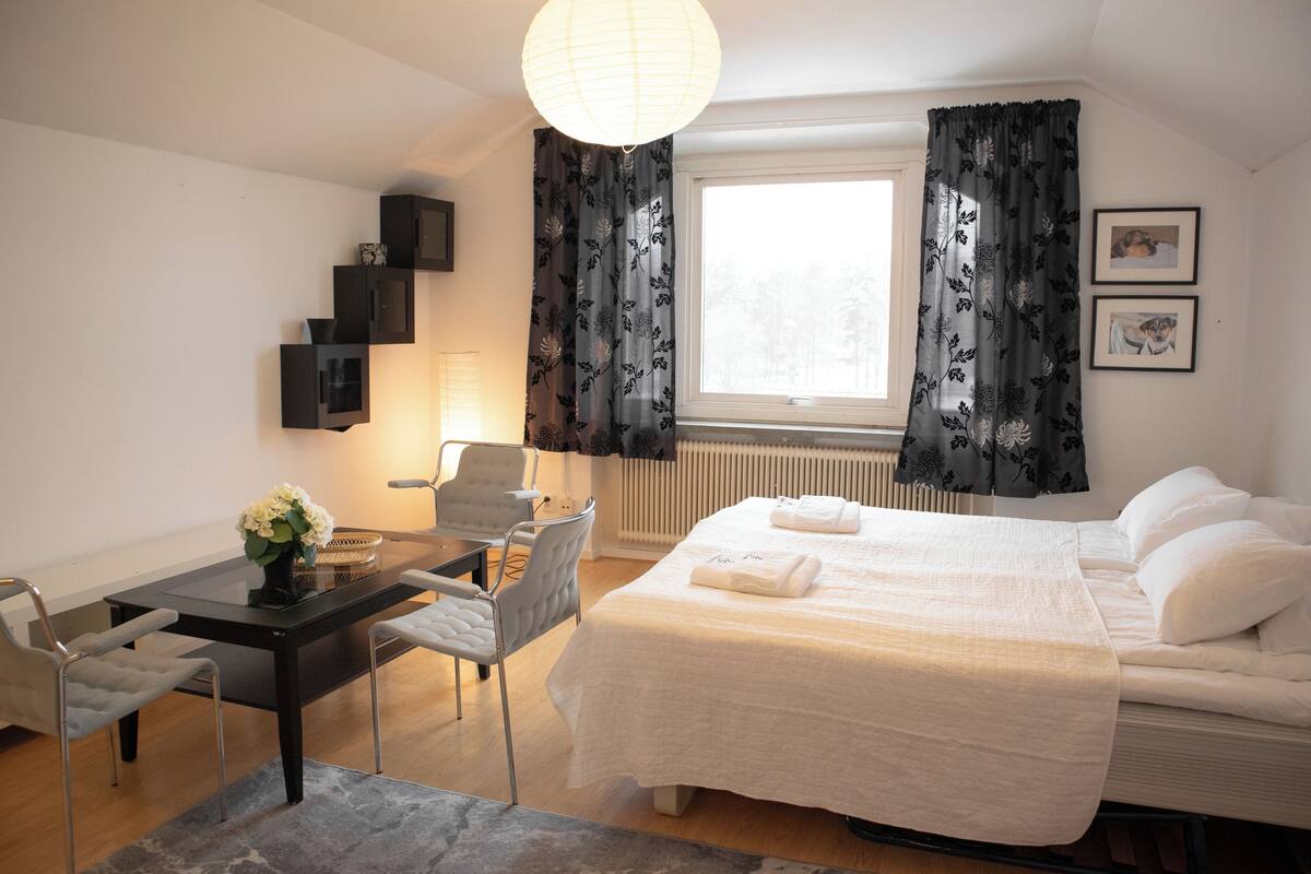Quarto de hotel com cama, escrivaninha com cadeira, luminária e janela com cortinas.
