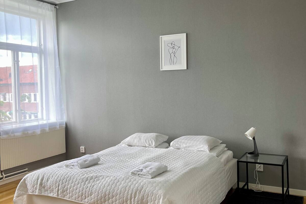 Quarto com cama, mesa de cabeceira com luminária, quadro na parede e janela com cortinas.