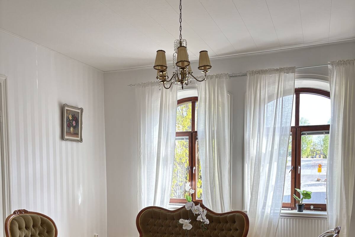 Um quarto com uma mesa redonda, duas poltronas, um candelabro e uma janela grande com cortinas.