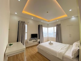 Quarto de hotel moderno com uma cama grande, televisão em prateleira embutida, uma mesinha com uma planta e vista da cidade através de grandes janelas.