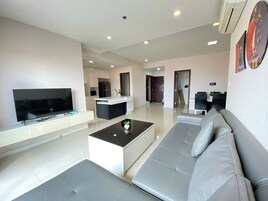 Uma sala de estar moderna com uma televisão de tela plana grande, um sofá cinza, uma mesa de centro e uma cozinha ao fundo.
