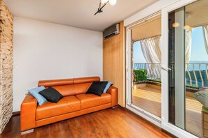 Quarto com piso de madeira, sofá de couro com duas almofadas, parede de pedra e porta de vidro deslizante que dá para uma varanda externa com guarda-corpo.