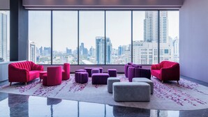 Quarto de hotel moderno com grandes janelas, vista para a cidade e mobiliário em tons de vermelho e roxo.