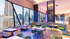 Restaurante moderno com vista para a cidade, com mesas altas, cadeiras coloridas e área de sofás verdes.