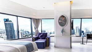 Quarto de hotel moderno com uma cama grande, área de estar com uma poltrona roxa, espelho redondo e vista para a cidade.