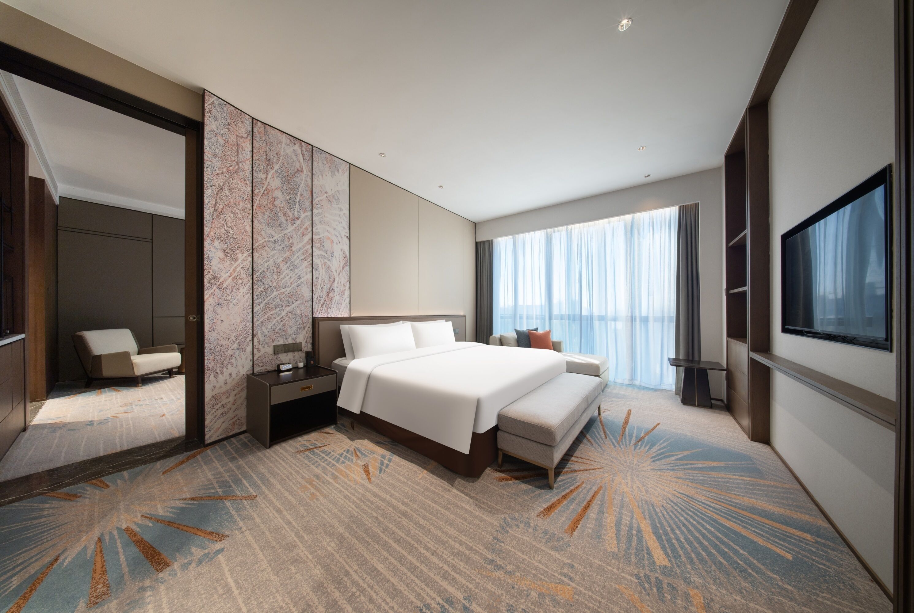 Quarto de hotel moderno com uma cama grande, área de estar, televisão e uma janela ampla com cortinas.