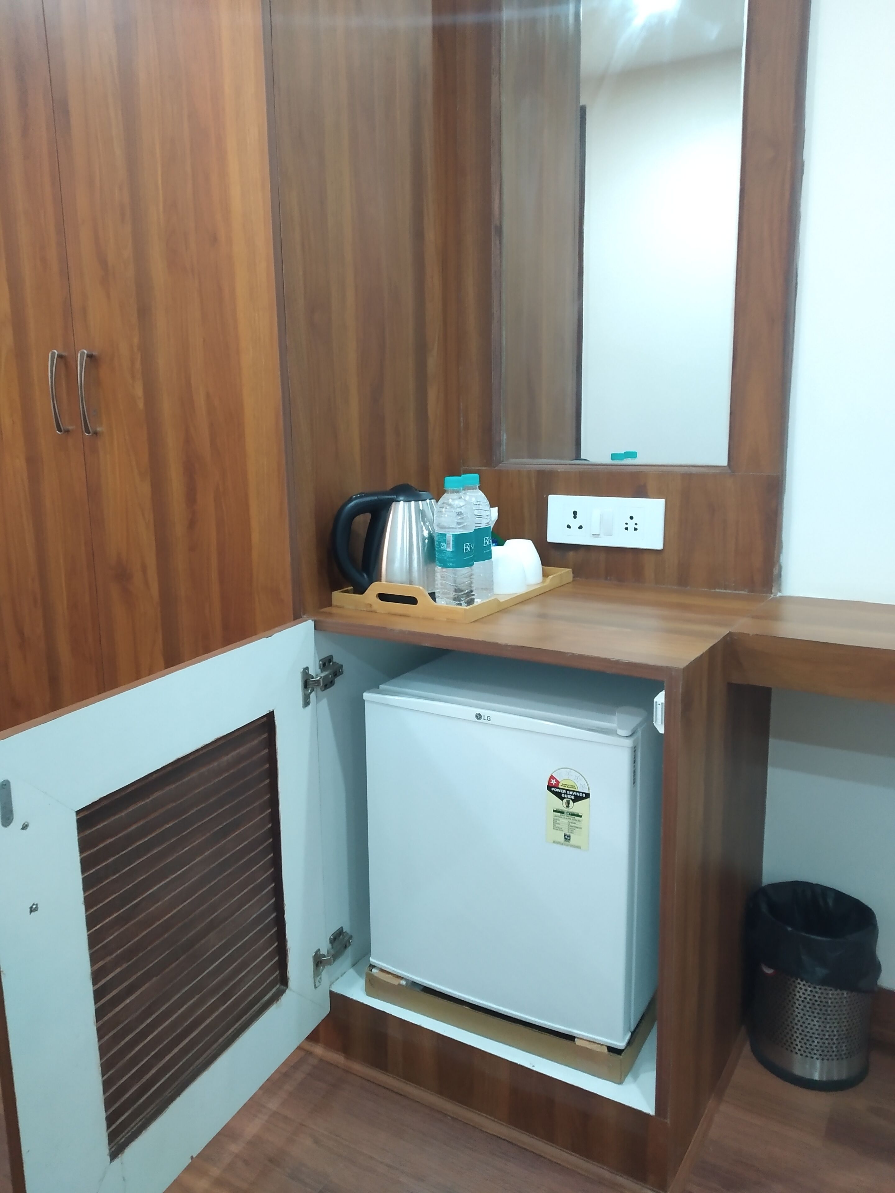 Quarto compacto com um guarda-roupa de madeira, uma pequena geladeira branca, uma prateleira de madeira com uma chaleira, garrafas de água e copos, e uma lixeira.