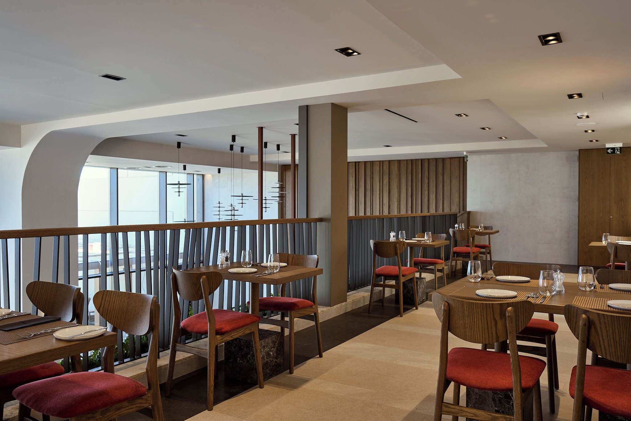 Restaurante moderno com mesas de madeira e cadeiras estofadas vermelhas, oferecendo vista da cidade por grandes janelas.